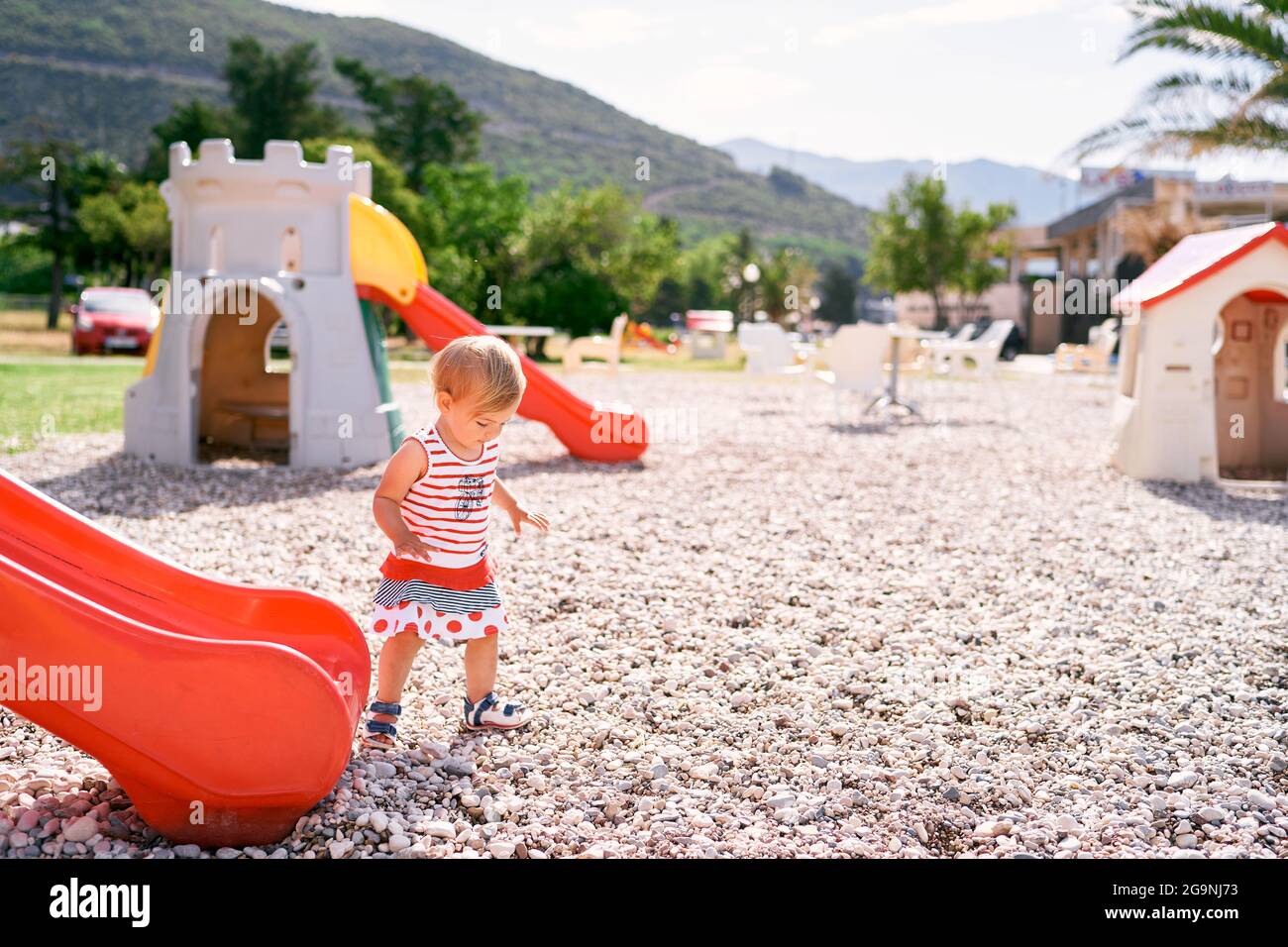 Little girl in a dress stands on a pebble playground near the slide ...