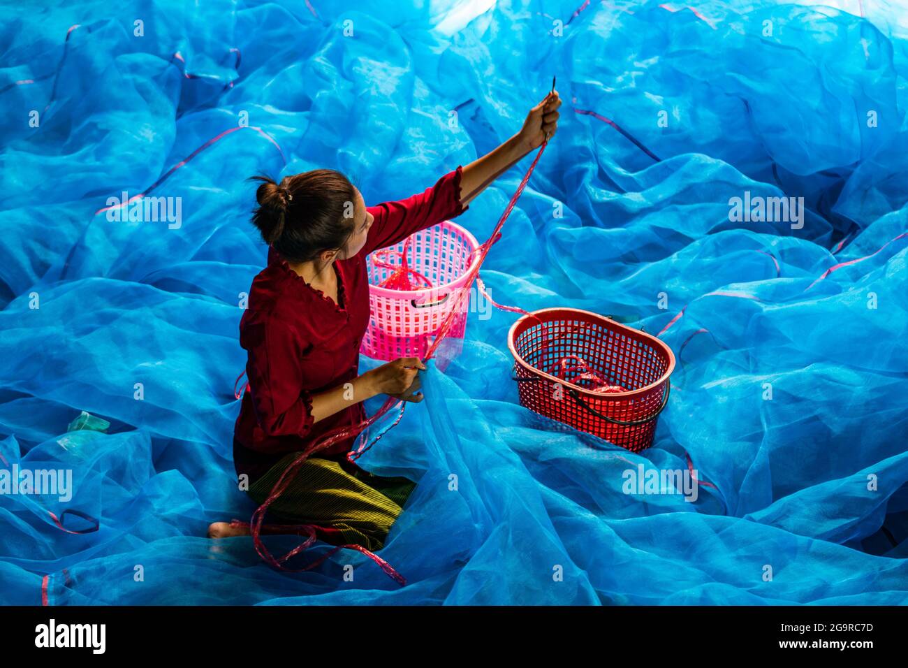 Overhead shot of a Southeast Asian woman sewing a blue fishnet as a ...