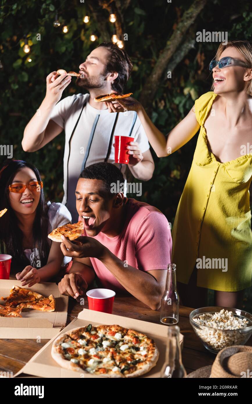 Positive interracial people eating tasty pizza near plastic cups ...