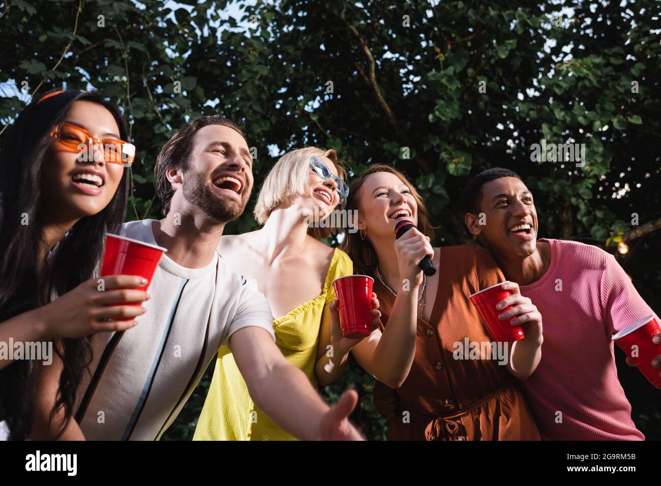 Cheerful multiethnic people singing karaoke and holding drink in ...