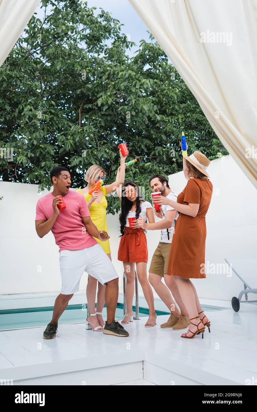 cheerful multiethnic friends with plastic cups and water pistols having ...