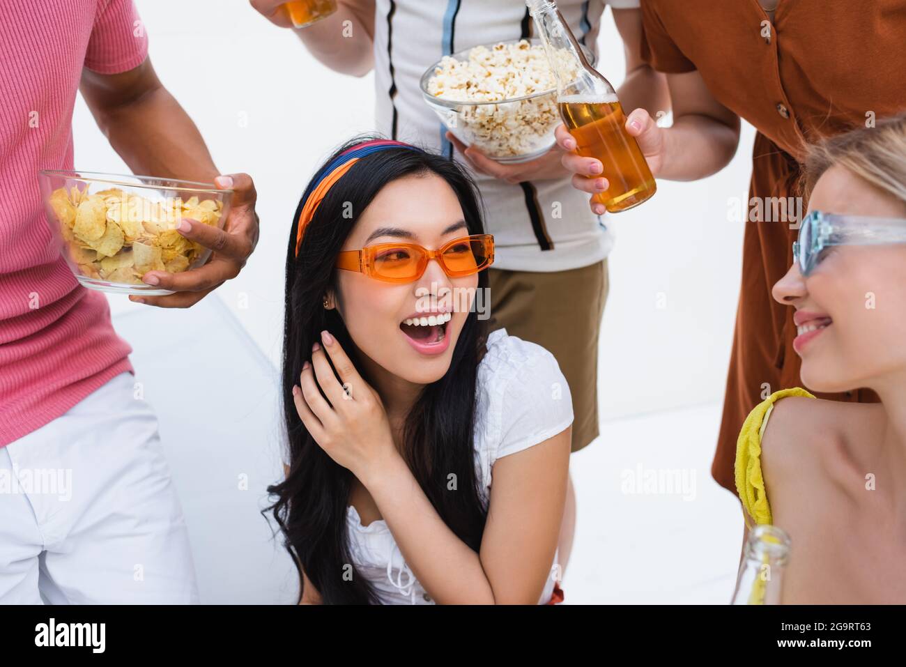 excited asian woman in sunglasses laughing near friends with snacks and ...