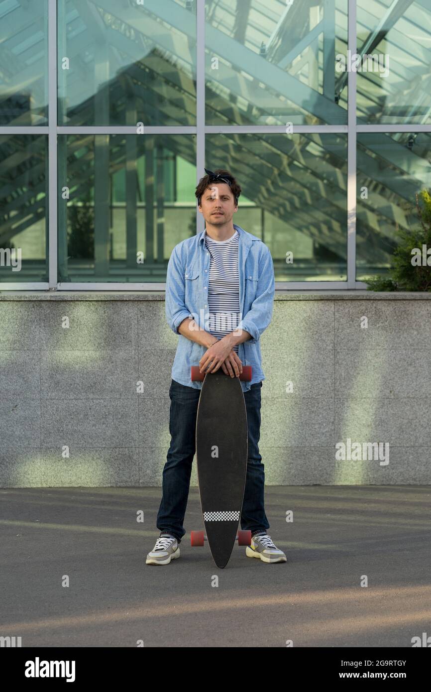Stylish man holding longboard over city building. Hipster guy ...