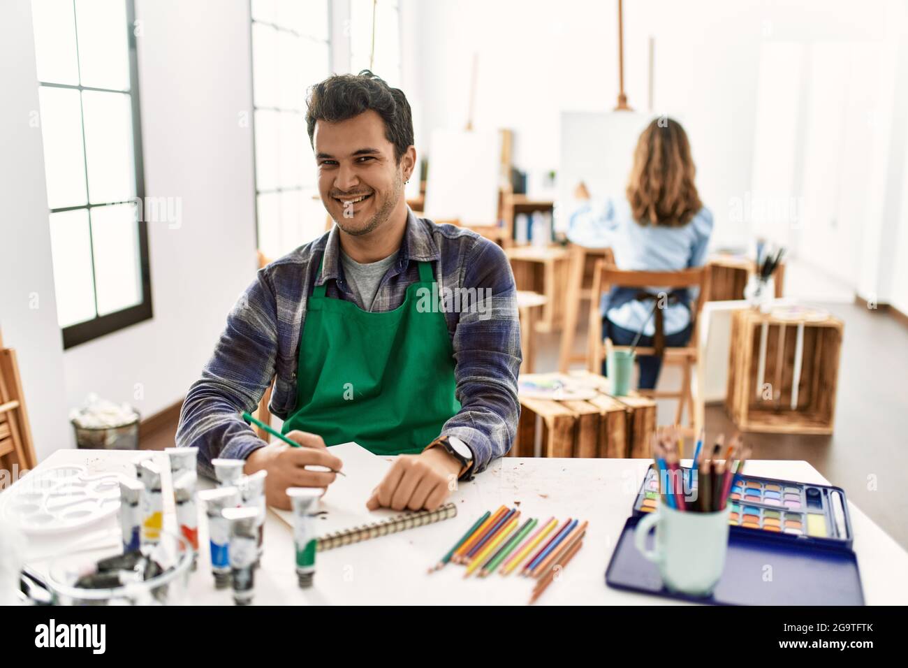 Young artist man at art studio looking away to side with smile on face ...
