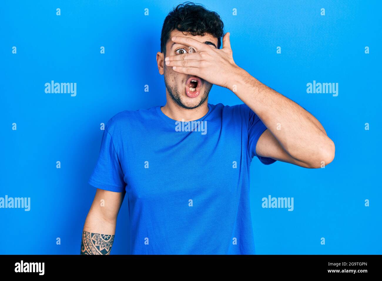 Young hispanic man wearing casual blue t shirt peeking in shock ...