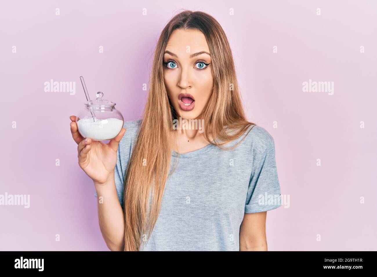 Young blonde girl holding bowl with sugar scared and amazed with open ...
