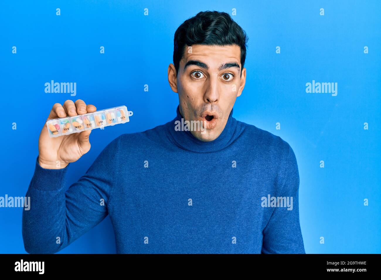 Handsome hispanic man holding pill organizer scared and amazed with ...