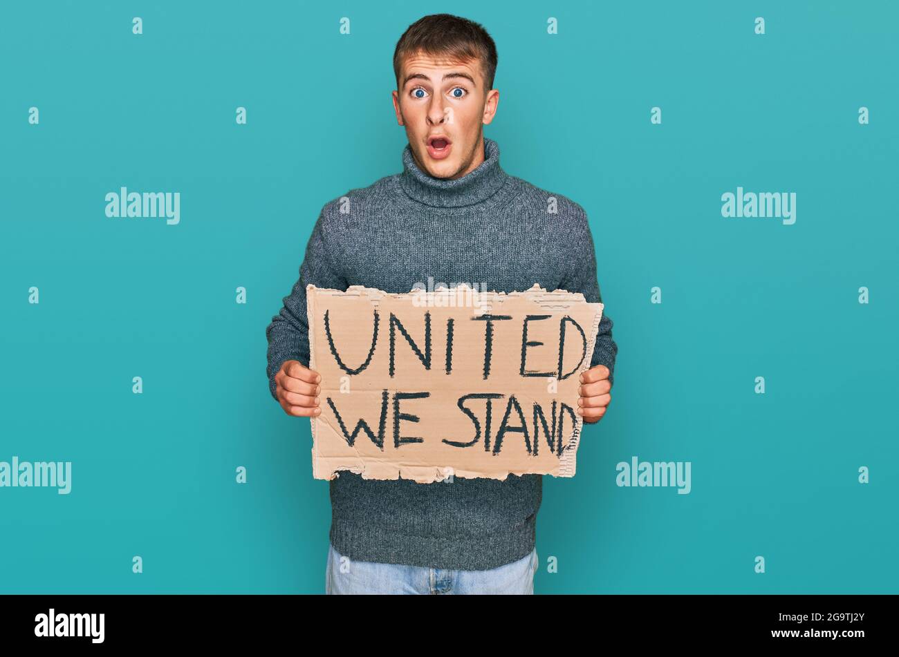 Young blond man holding united we stand banner scared and amazed with ...