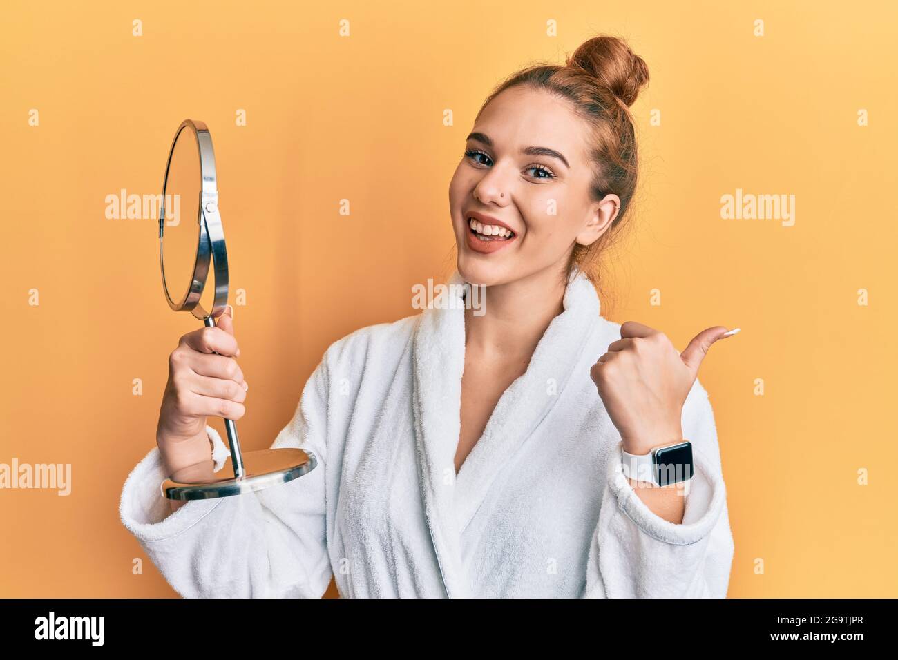 Young blonde woman wearing robe holding mirror pointing thumb up to the ...