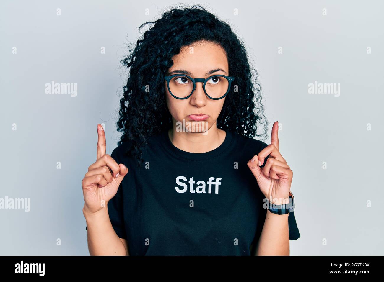 Young hispanic woman with curly hair wearing staff t shirt pointing up ...