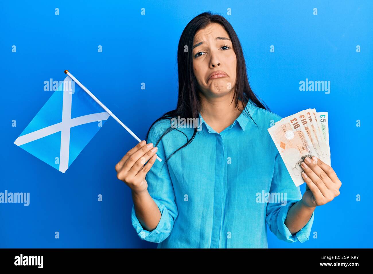 Young latin woman holding scotland flag and pounds banknotes depressed ...