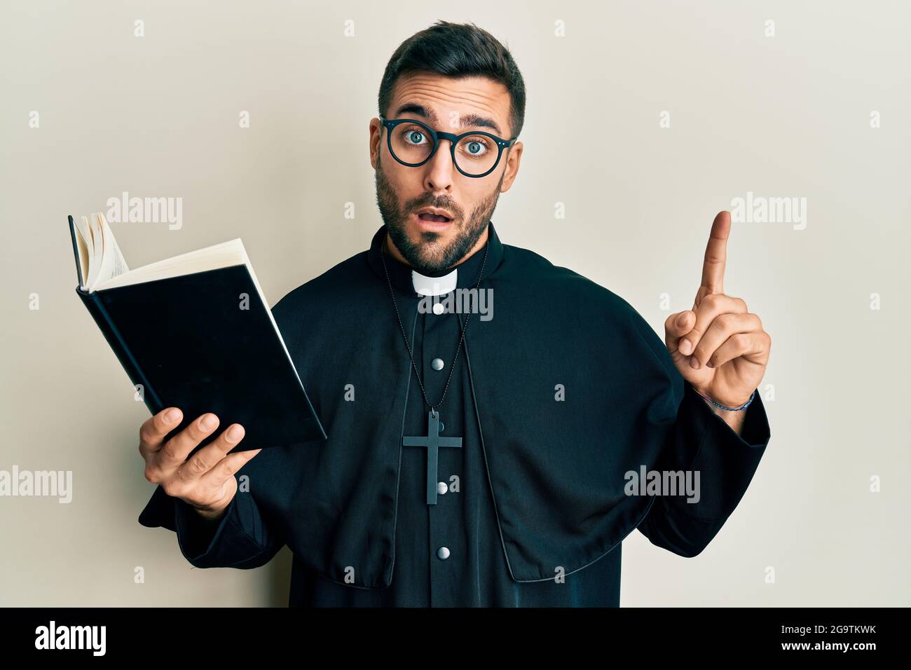 Young hispanic priest man holding bible with finger up in shock face ...
