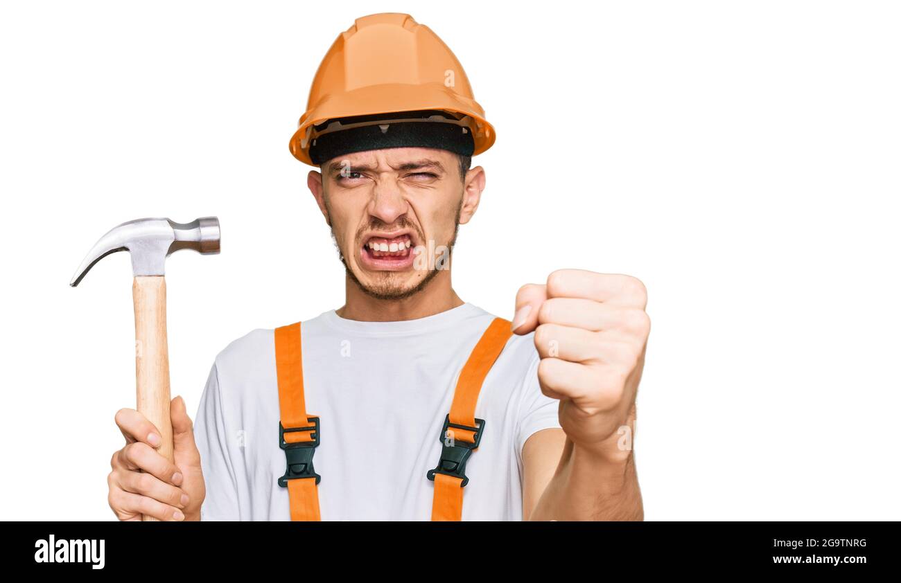 Hispanic young man wearing hardhat holding hammer annoyed and ...