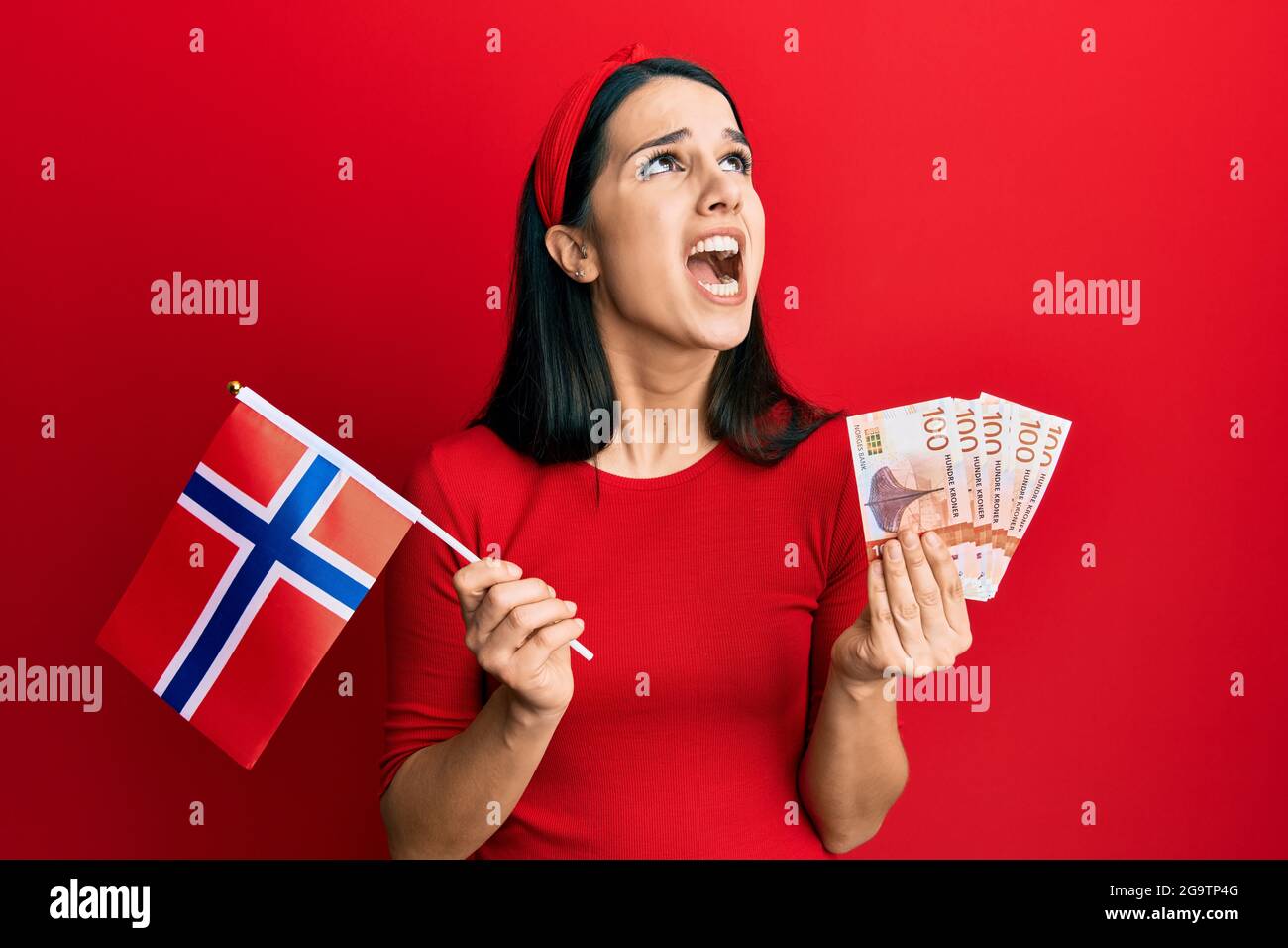 Young hispanic woman holding norway flag and krone banknotes angry and ...
