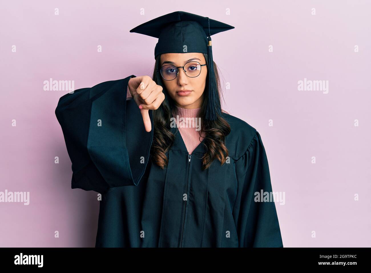 Young hispanic woman wearing graduation cap and ceremony robe looking ...