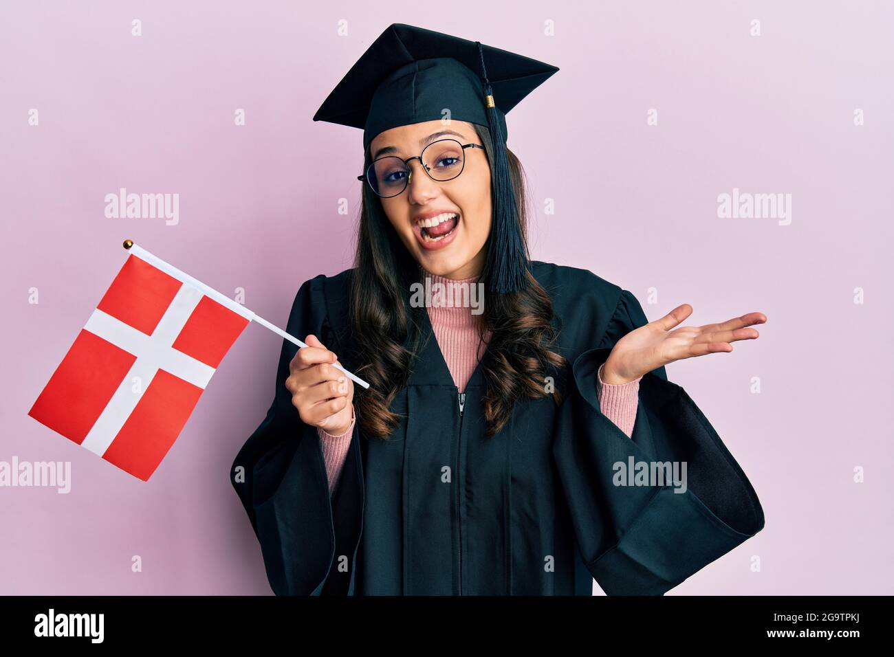 Young hispanic woman wearing graduation uniform holding denmark flag ...
