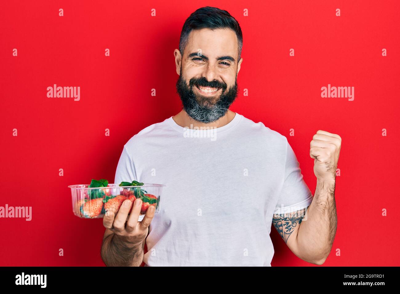 Young hispanic man holding strawberries screaming proud, celebrating ...