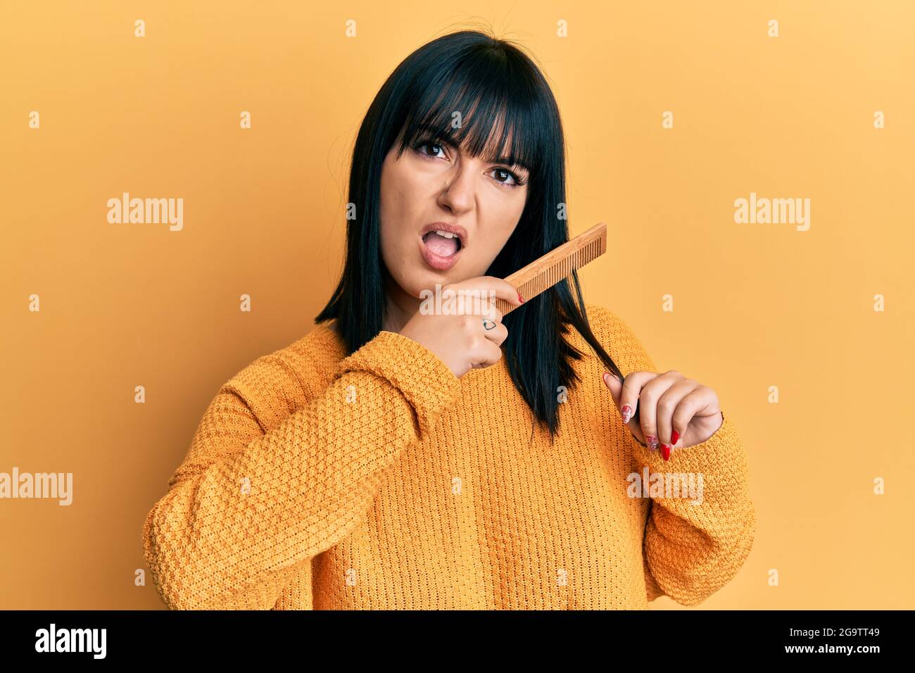 Young hispanic woman styling hair using comb in shock face, looking ...