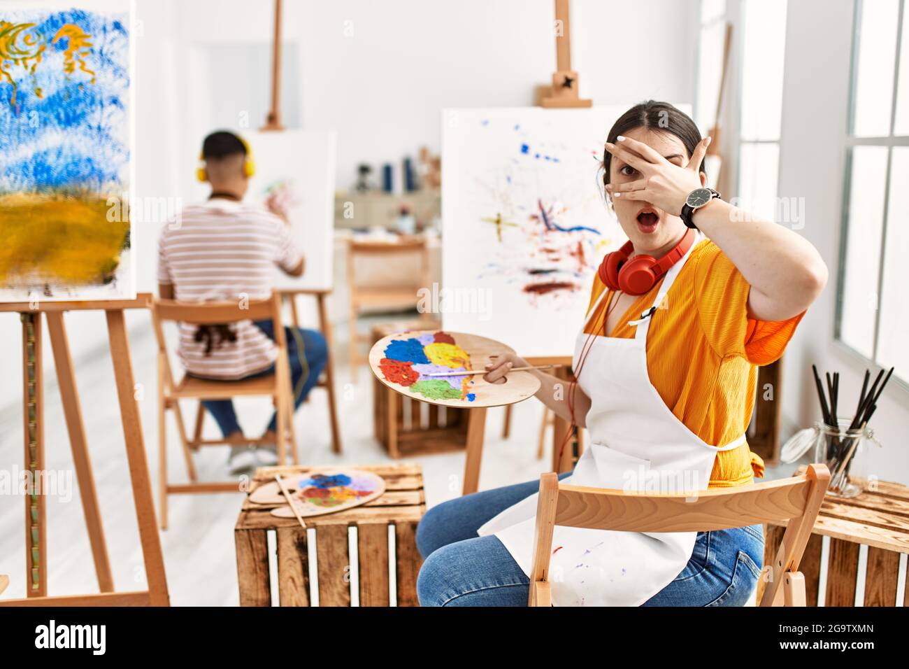 Young artist woman painting on canvas at art studio peeking in shock ...