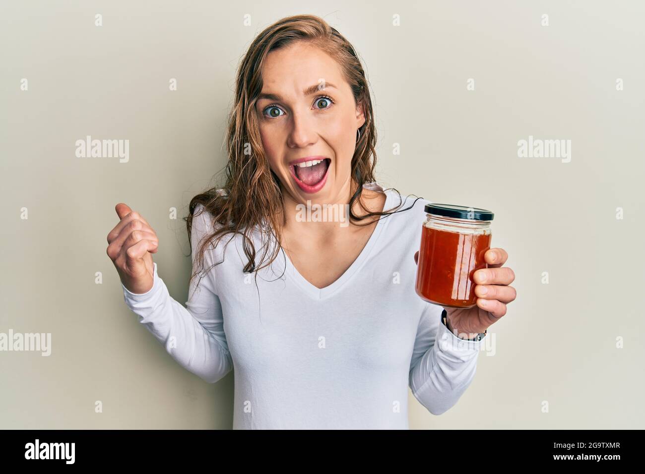 Young blonde woman holding jam pointing thumb up to the side smiling ...
