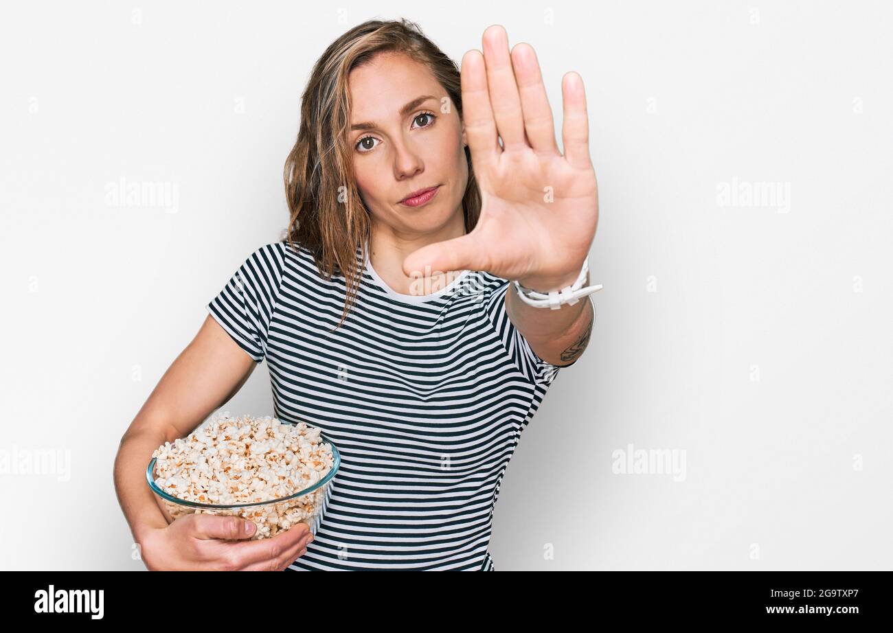 Young blonde woman eating popcorn with open hand doing stop sign with ...