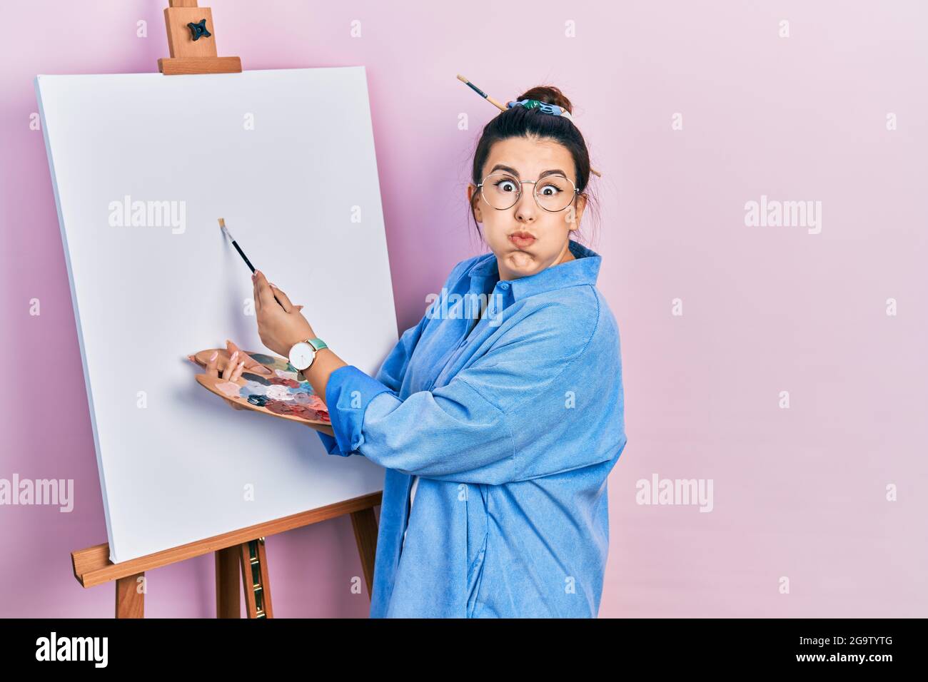 Young hispanic woman standing drawing with palette by painter easel ...