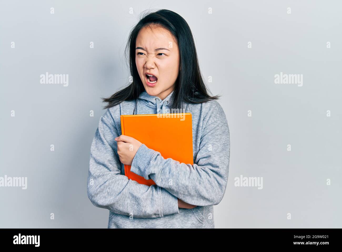 Young chinese girl holding book angry and mad screaming frustrated and ...