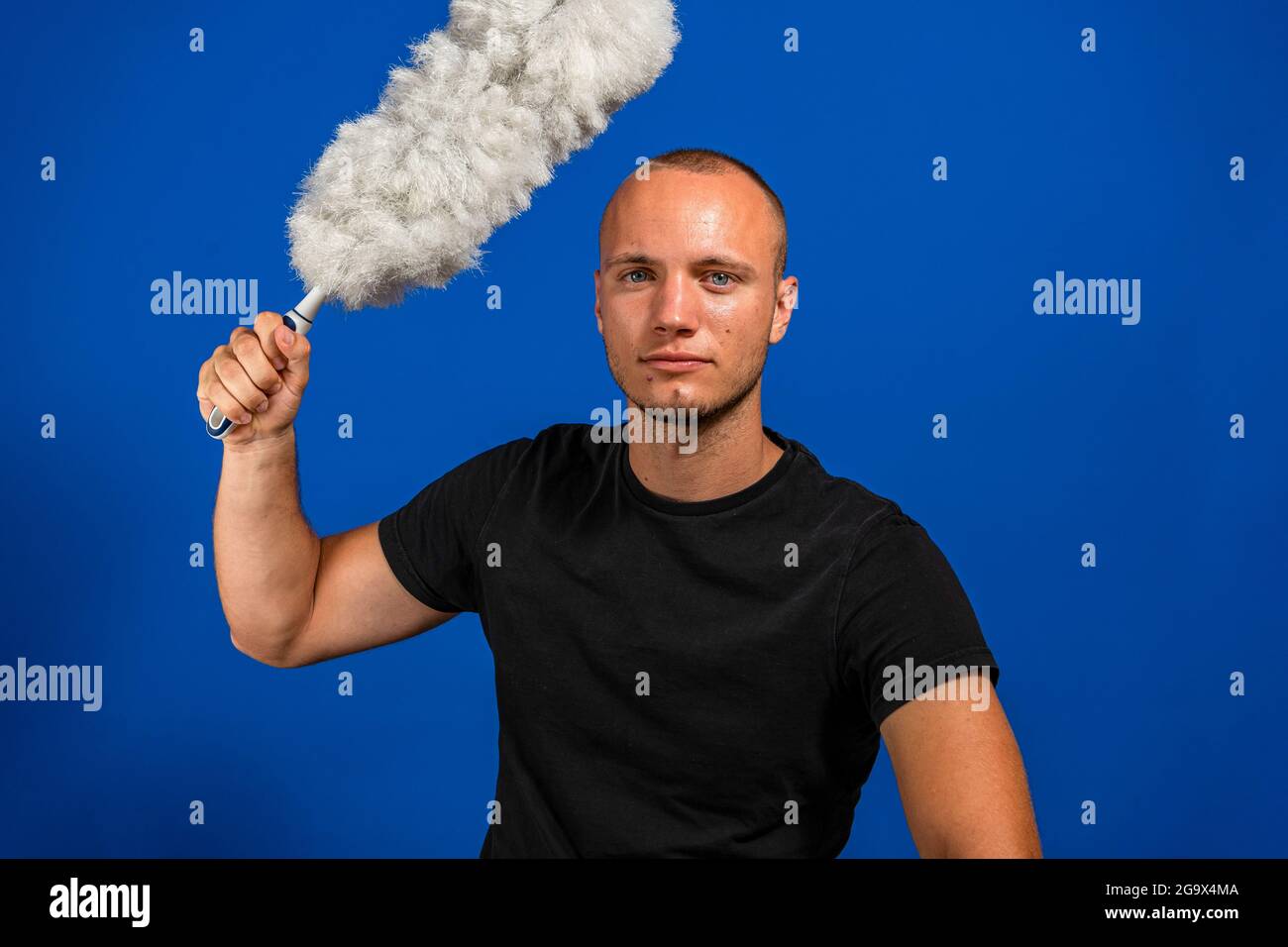 Caucasian young man posing funny with a cleaning duster on blue studio ...