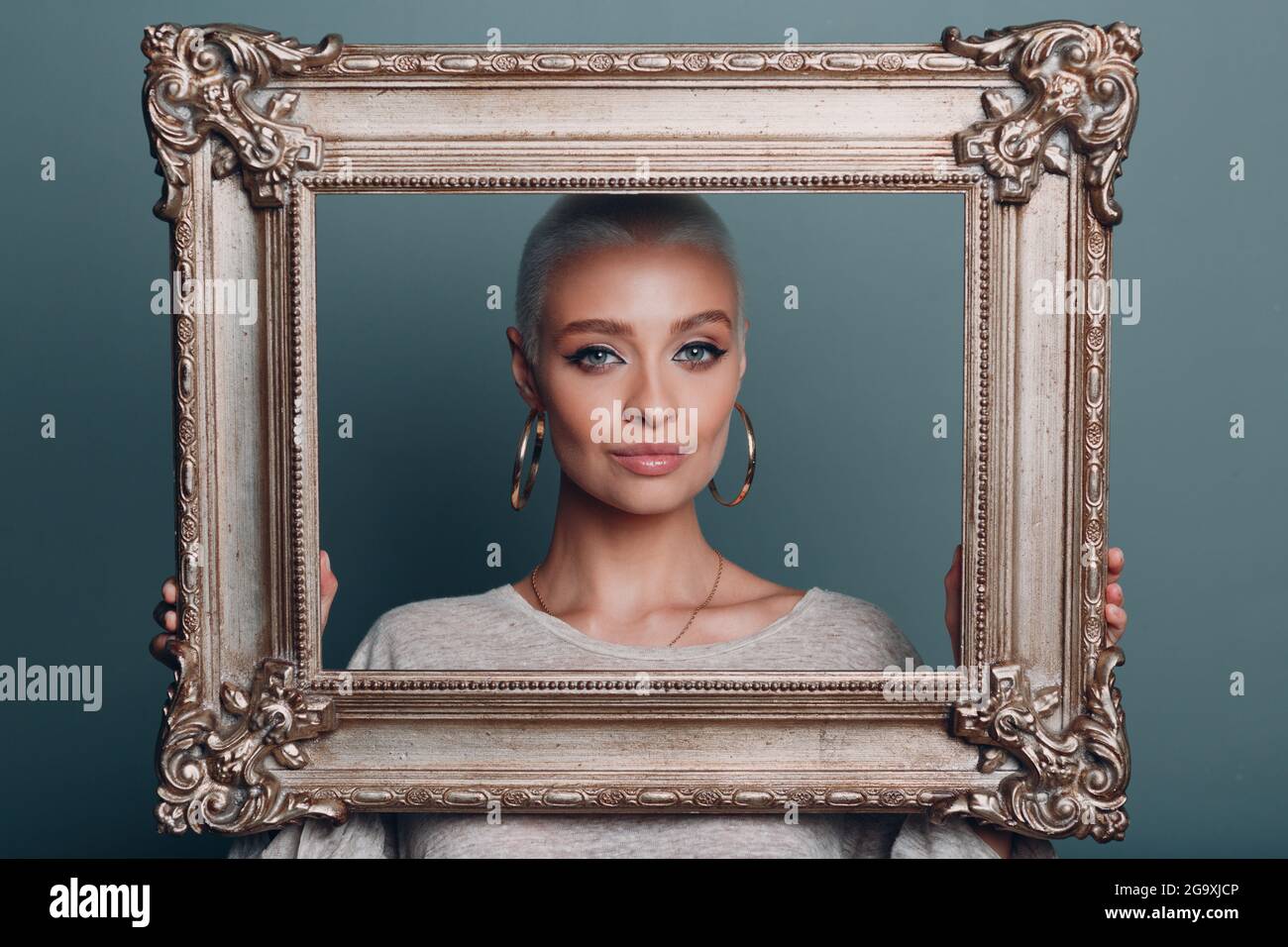 Millenial young woman with short blonde hair holds gilded picture frame ...