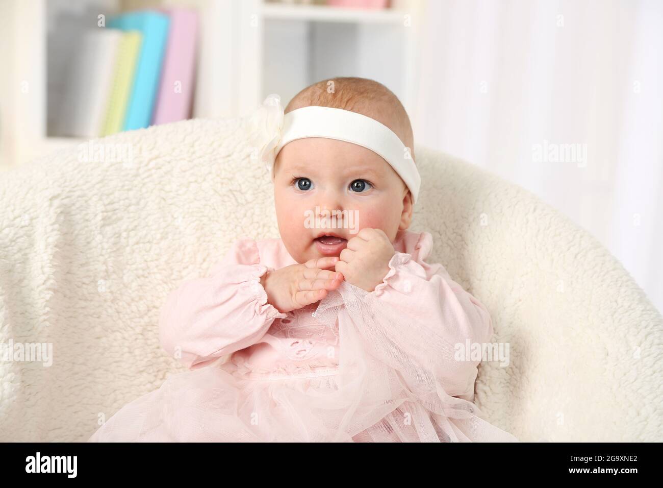 Cute baby girl in pink dress sitting in arm-chair, on home interior ...