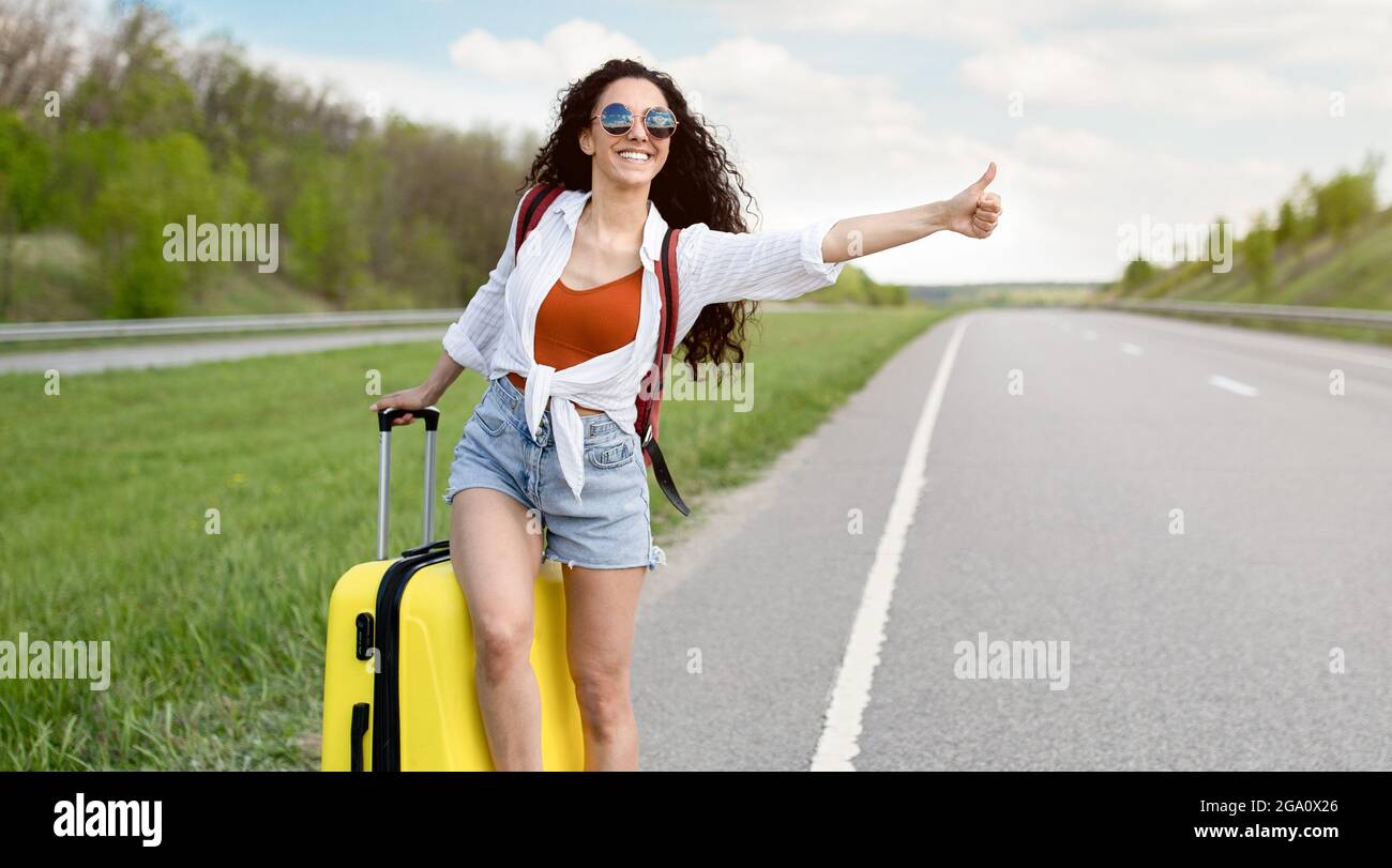 Young lady thumbing her ride, standing with suitcase on highway, trying ...
