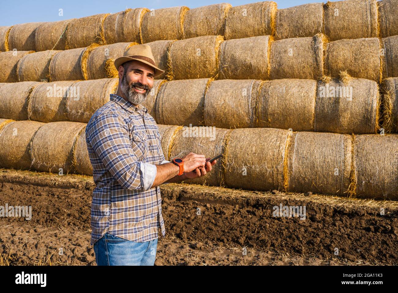 Happy farmer is standing beside bales of hay. He is examining straw ...