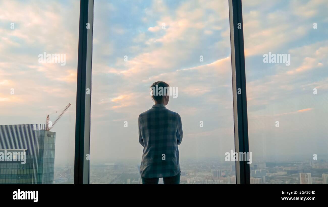 Pensive woman looking at cityscape through window of skyscraper - back ...