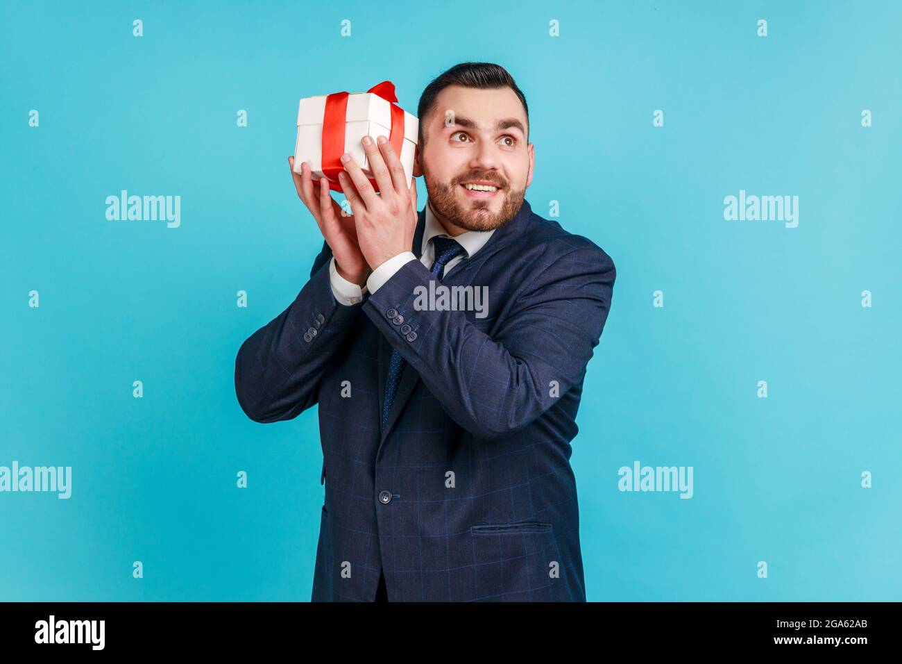Curious dreamy bearded man in elegant suit shaking gift box trying to ...