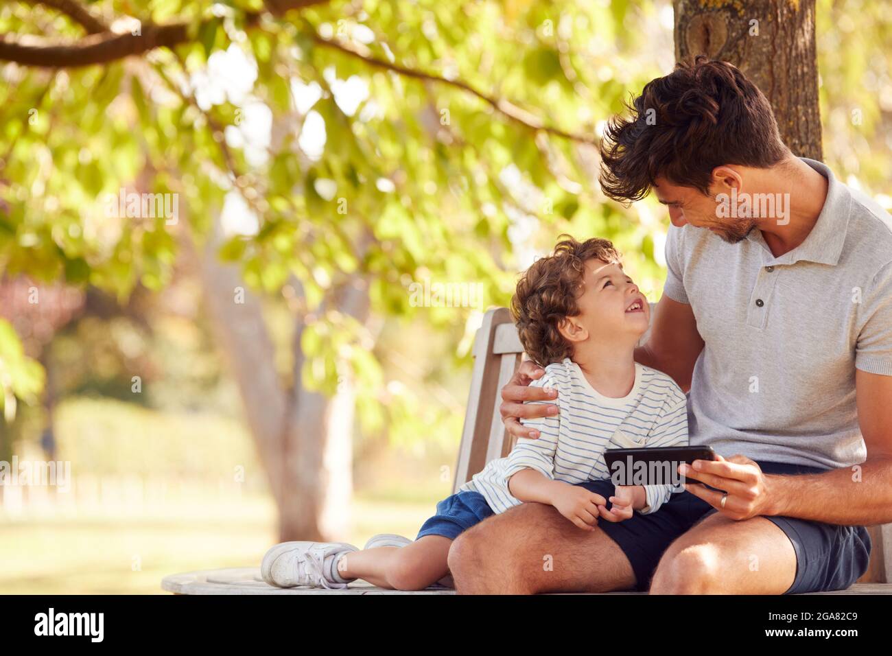 Father Sitting On Park Bench Under Tree With Son Looking At Mobile