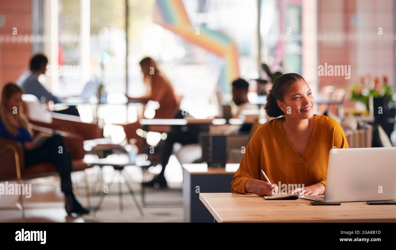 Young Businesswoman Sitting At Desk Writing In Notebook In Modern Open ...