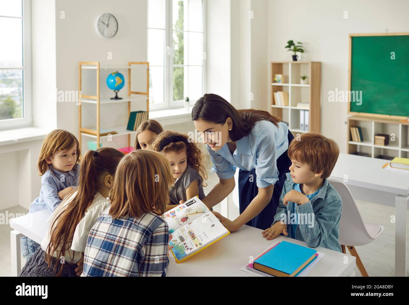 Young friendly female teacher in the classroom with a book in her hands teaches young students ...