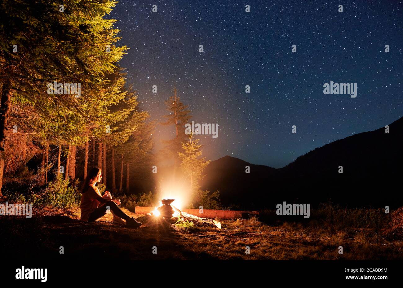 Young woman tourist sitting on bench near campfire under magical sky ...