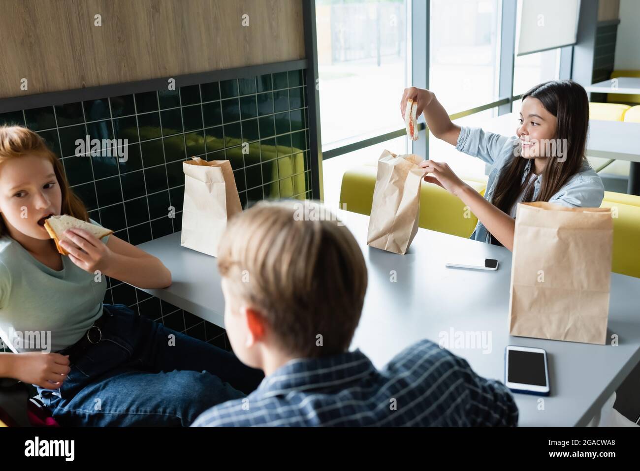 happy teenage girl holding sandwich near classmates in school canteen ...