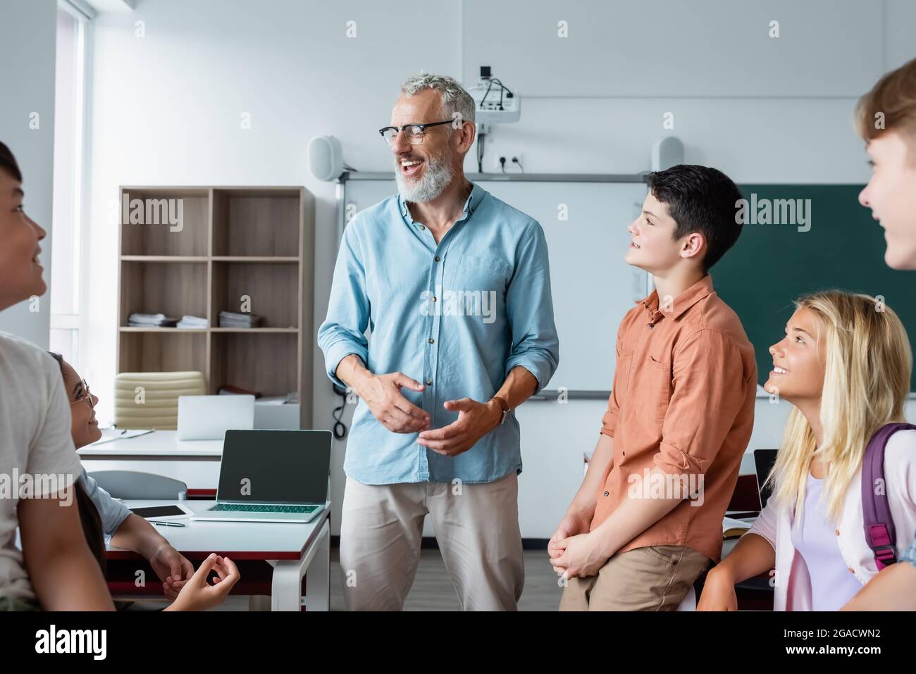Cheerful teacher talking to pupils on blurred foreground in classroom ...