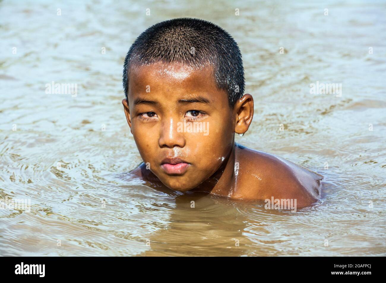 Close up of young Burmese boy with cropped hair bathing in murky water ...