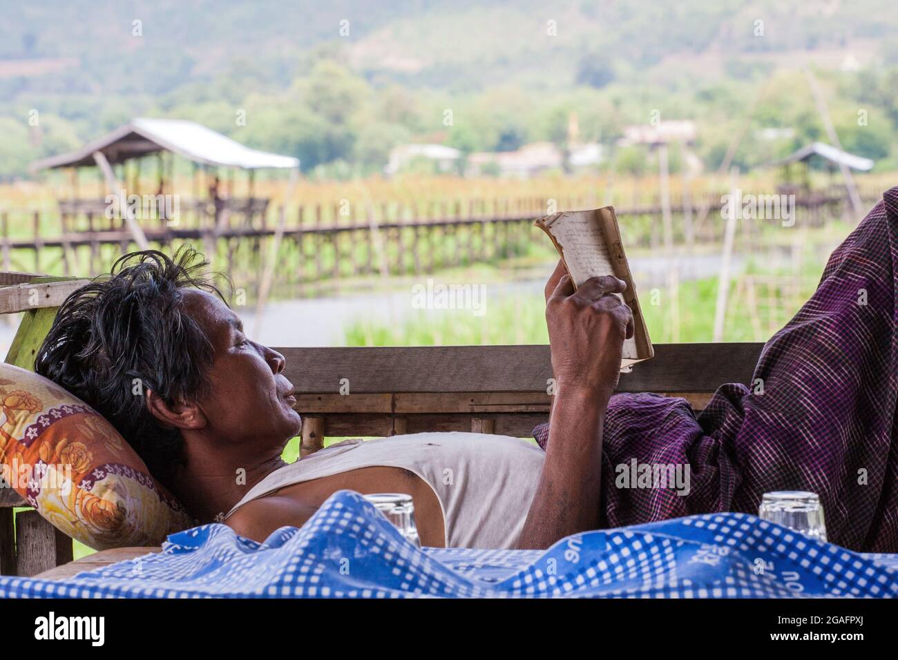 Middle aged Burmese male in vest and longyi reclines on couch reading ...