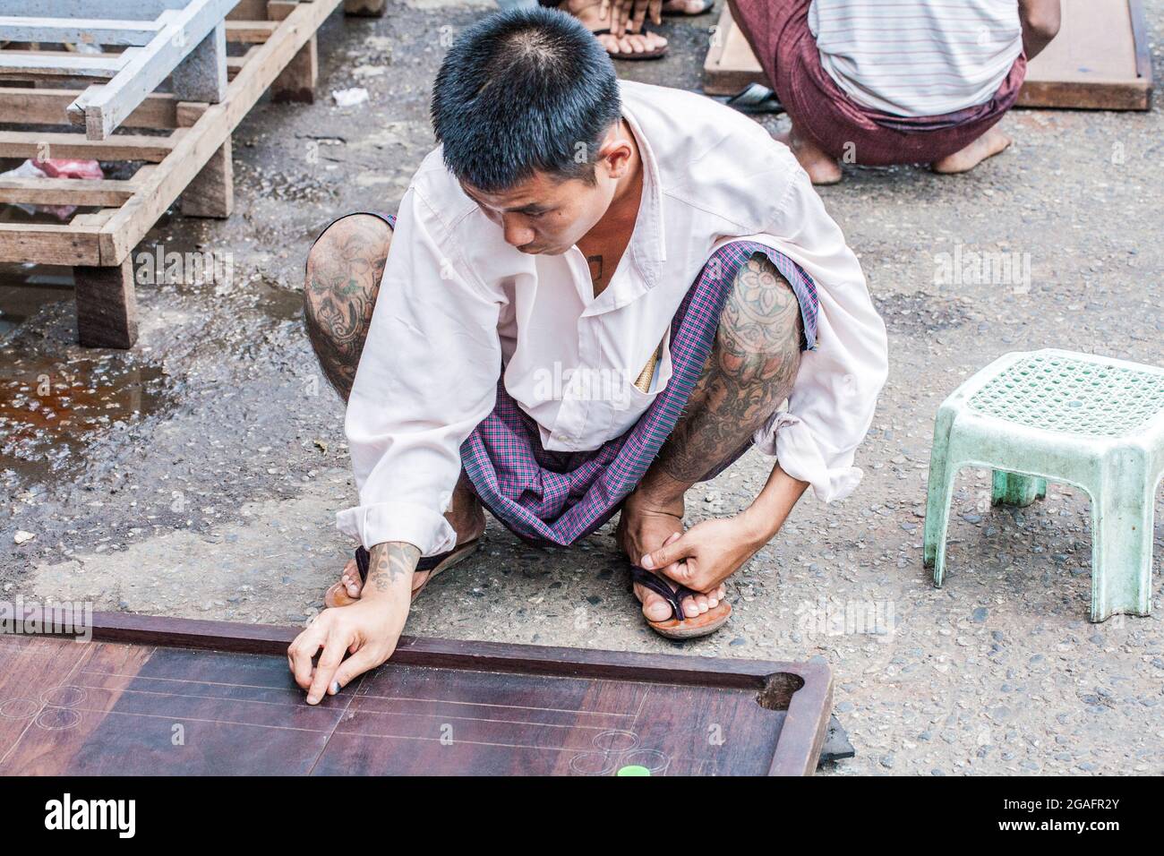 Burmese male wearing longyi with colourful leg tattoos playing billiards (carrom/karrom) on ...