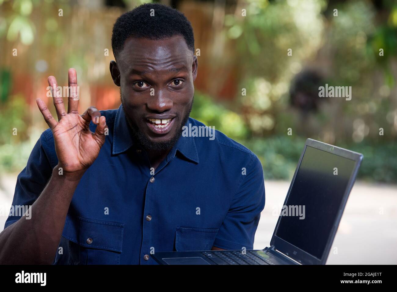 smiling young man sitting outdoors looking at camera and waving fingers ...