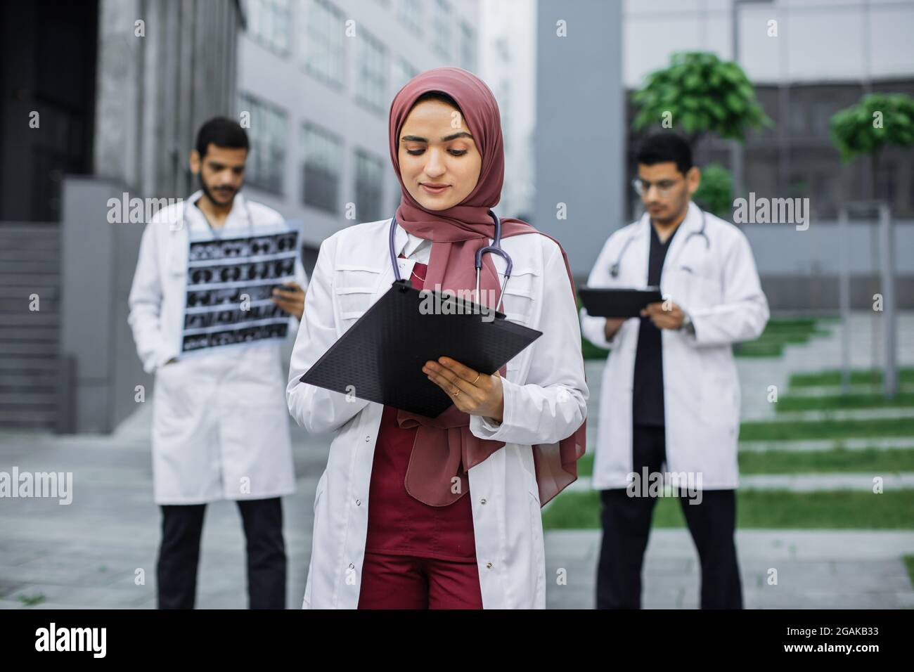 Pretty doctor in hijab writing notes on clipboard, standing ouside ...