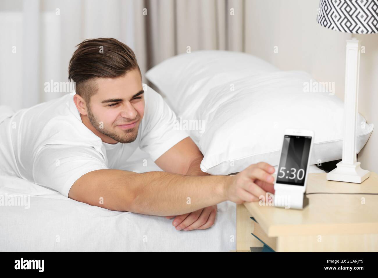 Beautiful young man waking up with mobile alarm clock Stock Photo - Alamy