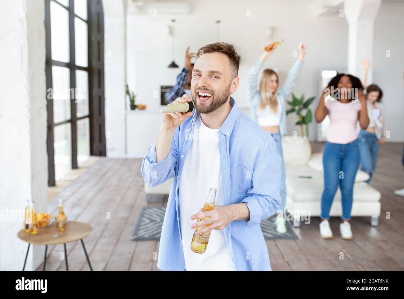 Attractive Caucasian guy with beer bottle singing karaoke on home party ...