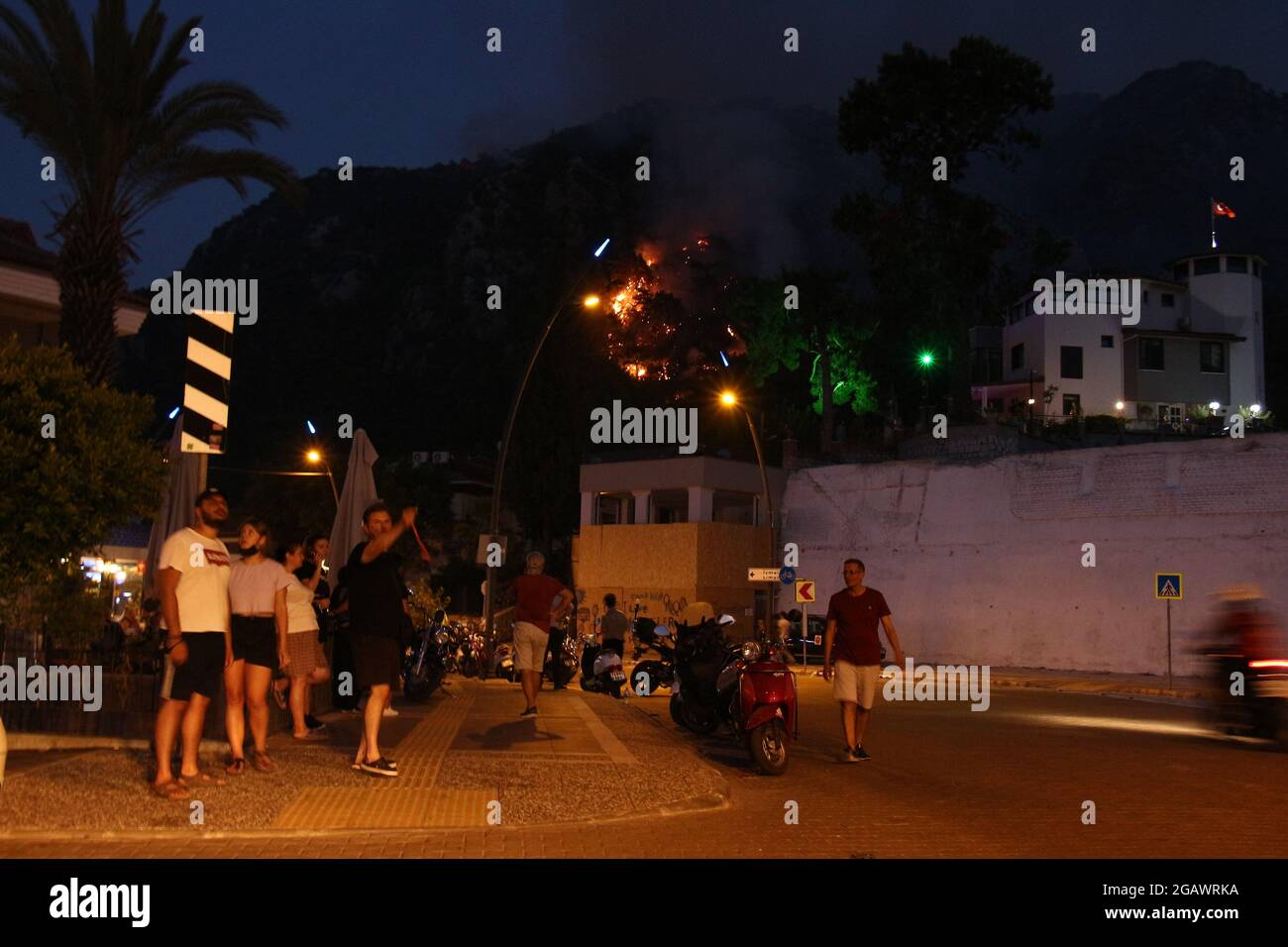People stand on a street as smoke and flames rise from a forest fire in ...