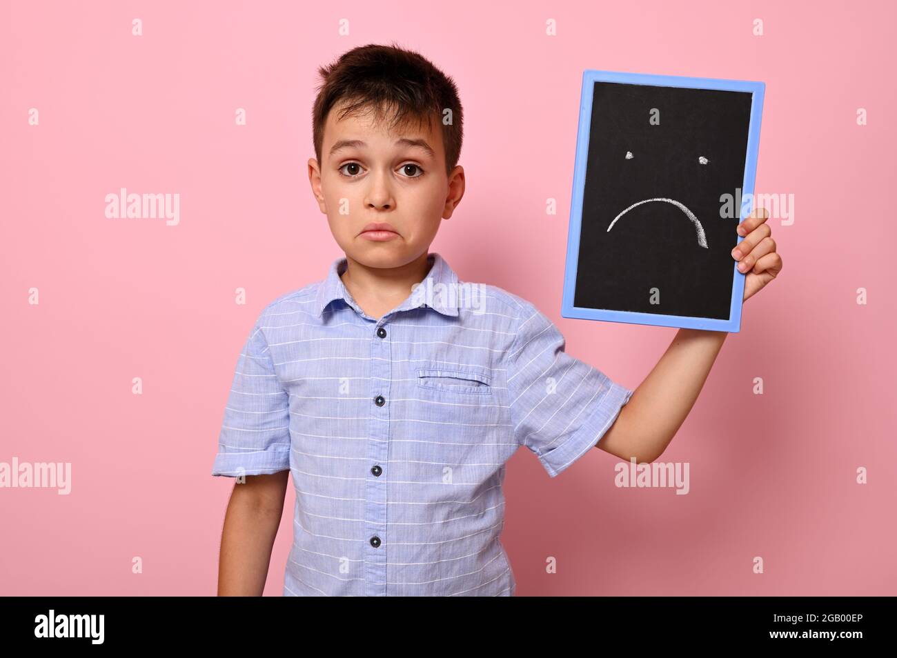 A frustrated boy in a blue shirt holds a blackboard near his face with ...