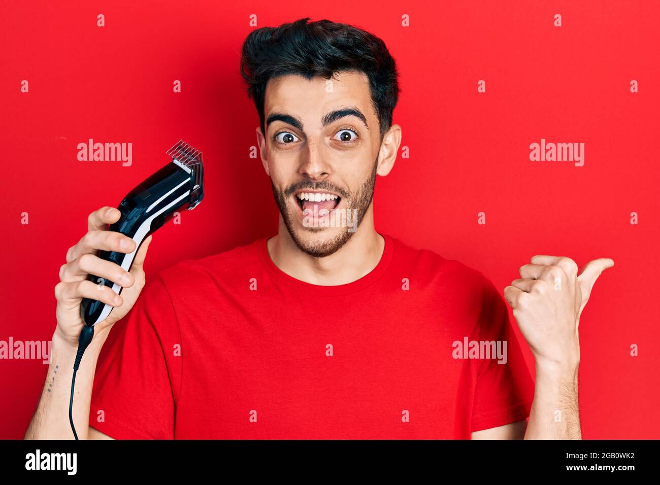 Young hispanic man charging electric razor machine pointing thumb up to ...
